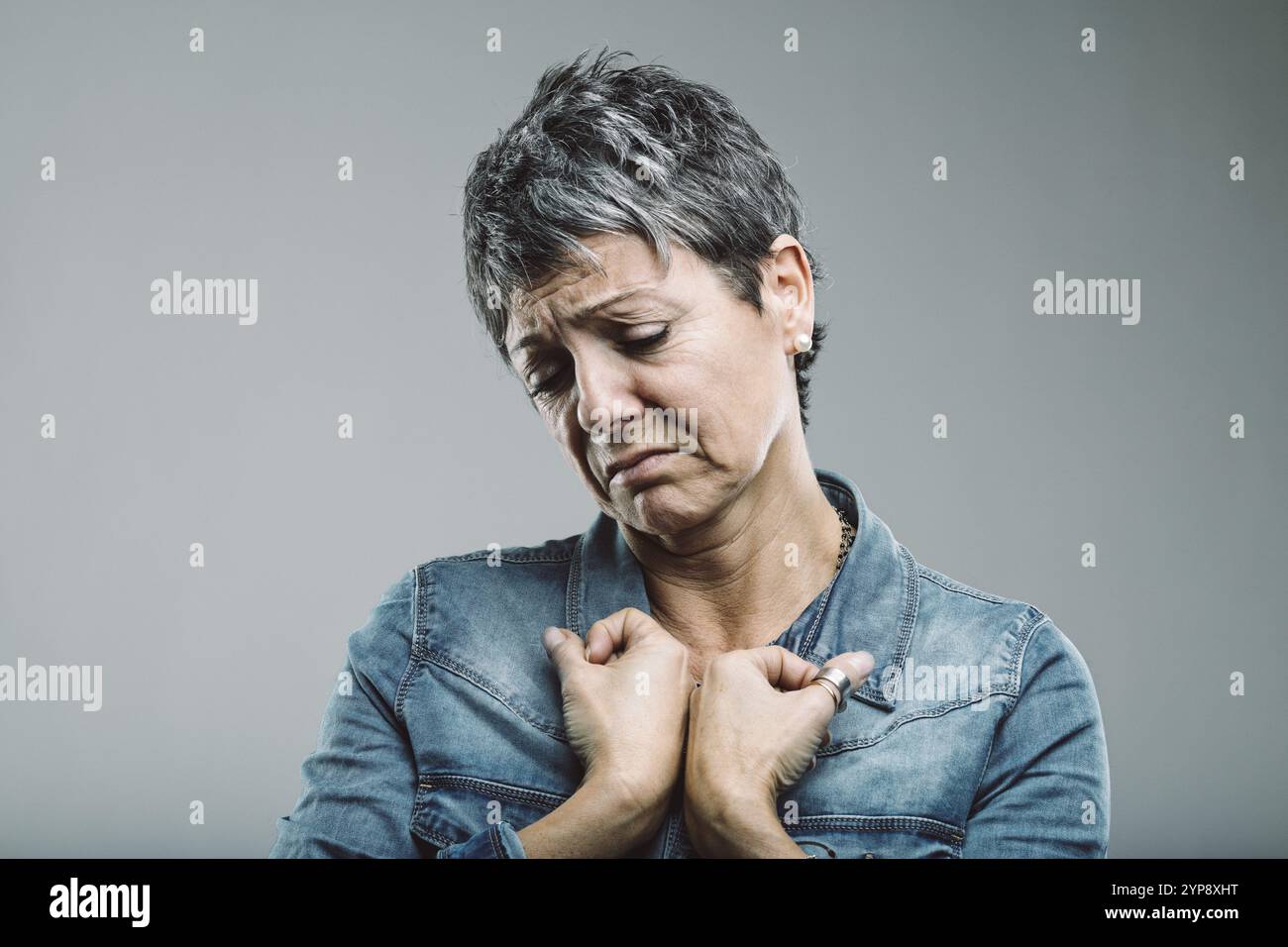 Mature woman with grey hair and denim shirt, looking down with a sad ...