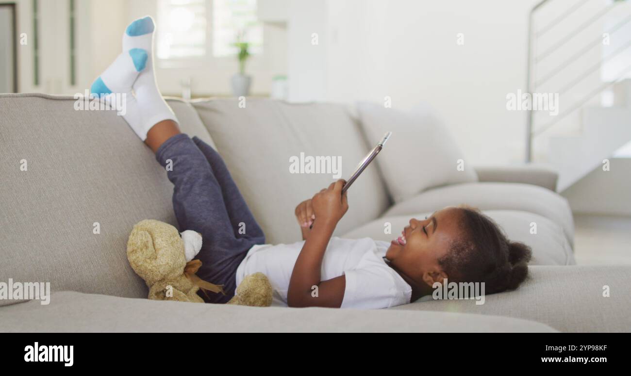 African american happy girl lying on sofa using digital tablet at home ...