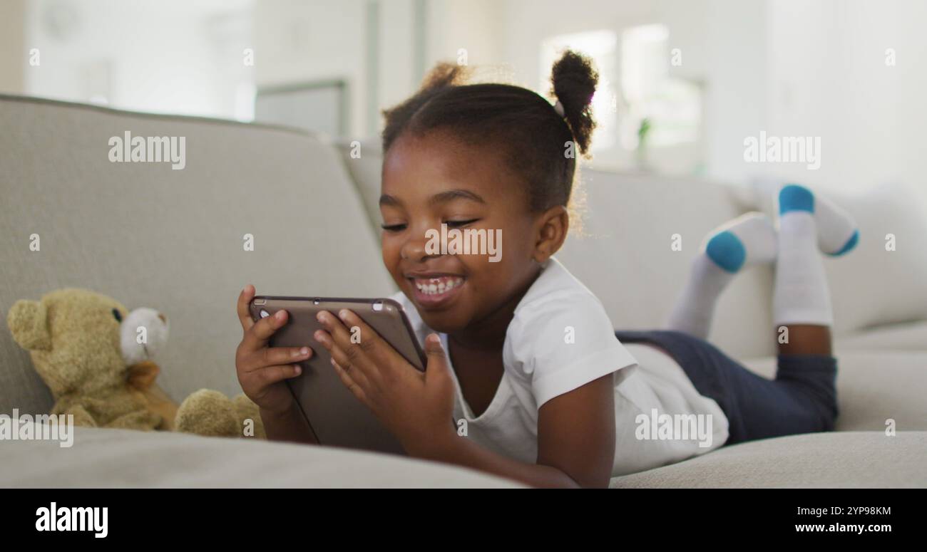 Happy african american girl lying on sofa using digital tablet at home ...