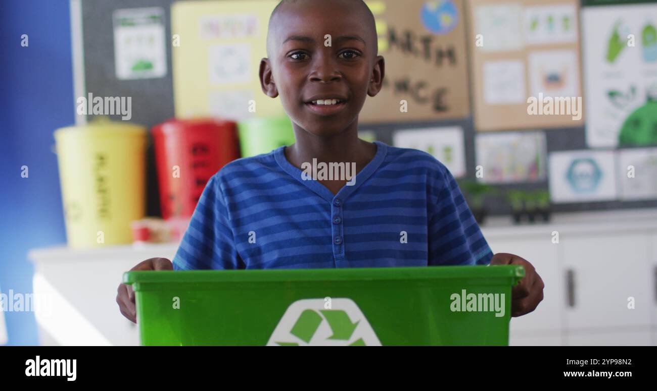 African american schoolboy smiling, holding recycling bin, standing in ...