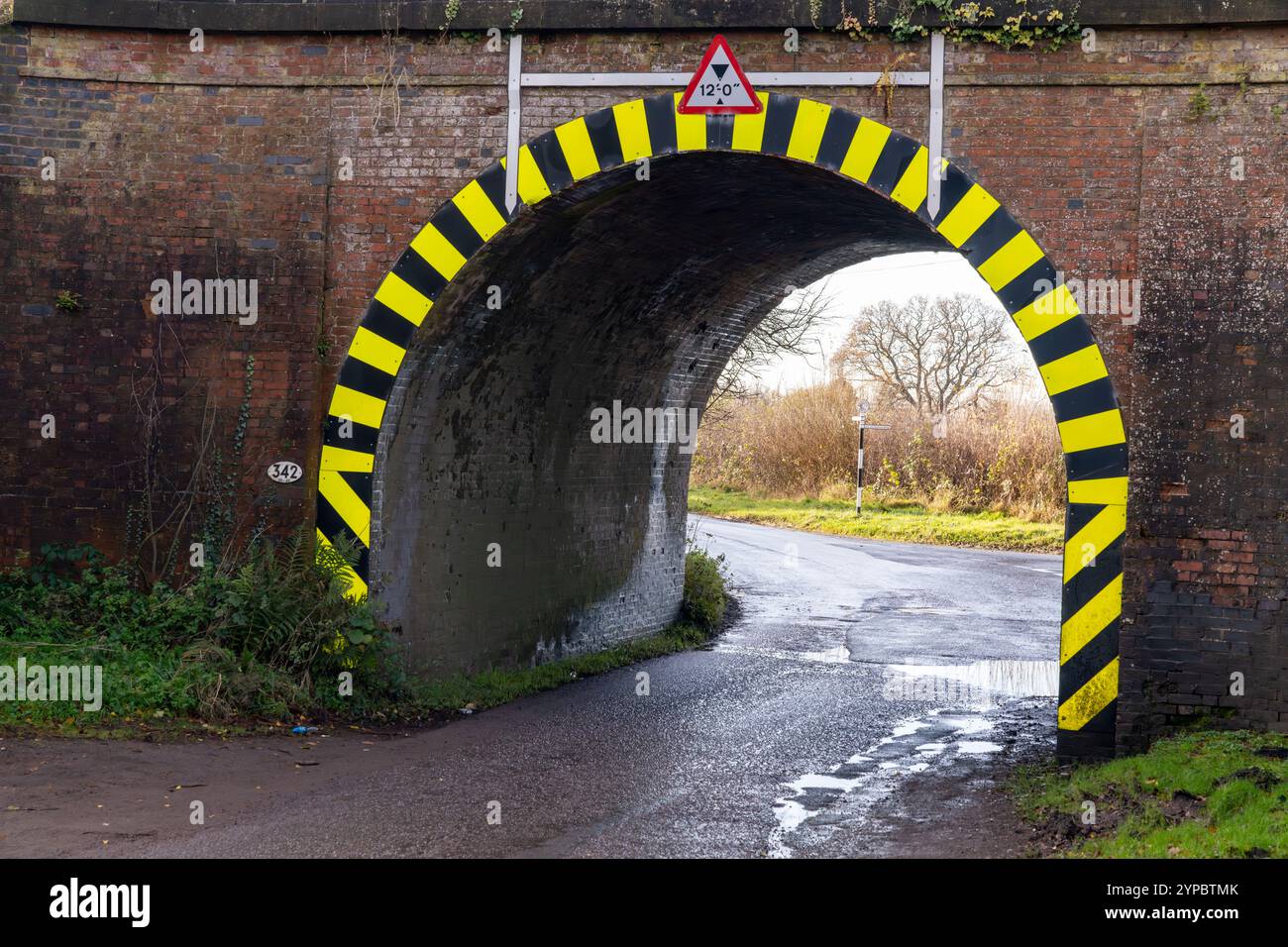 Narrow country road passing under a low railway bridge with black and ...