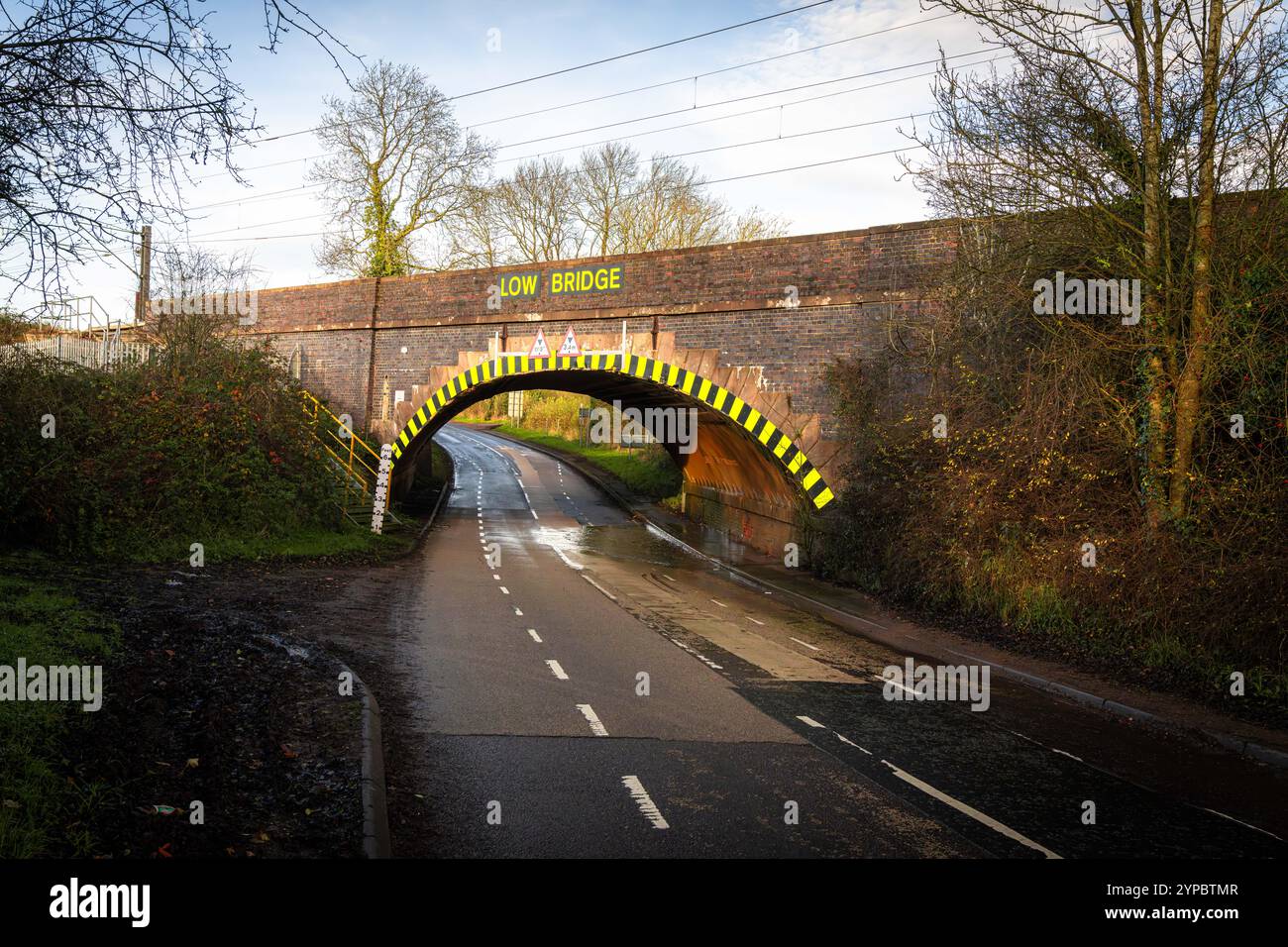 Wet road surface passing under a low railway bridge with black and ...