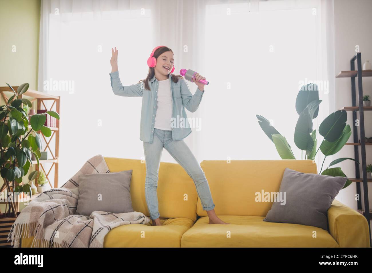 Joyful young girl singing with headphones and a microphone on a couch ...