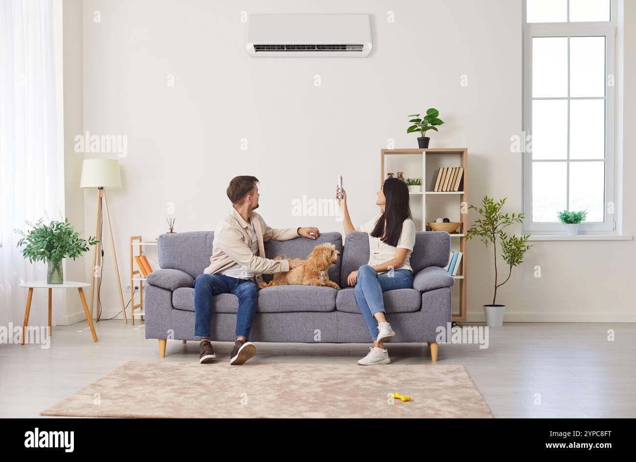 Young couple, happy family with dog on sofa switching on air conditioner, blowing warm, cold air ...