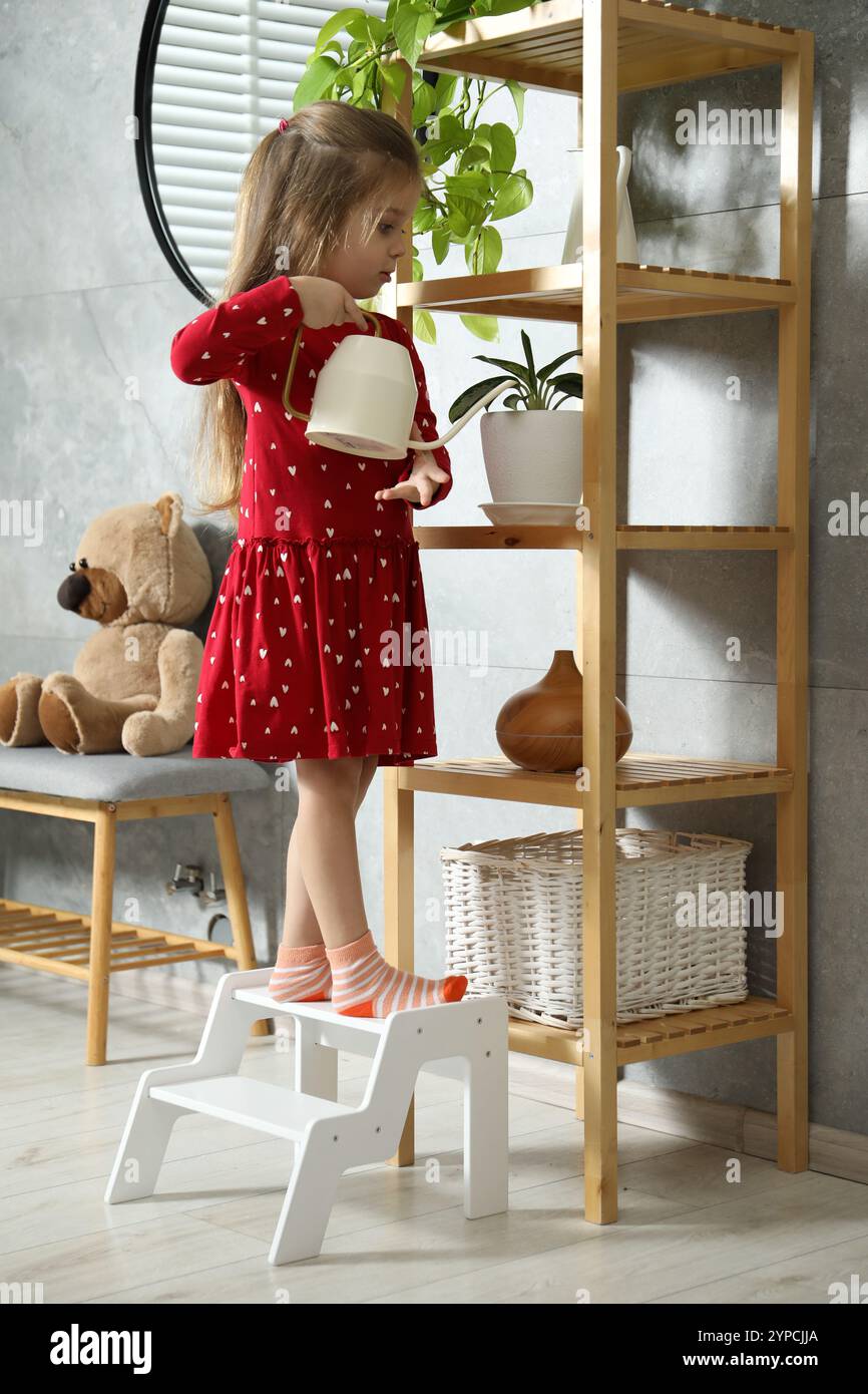 Little girl standing on step stool and watering plants on shelf indoors ...