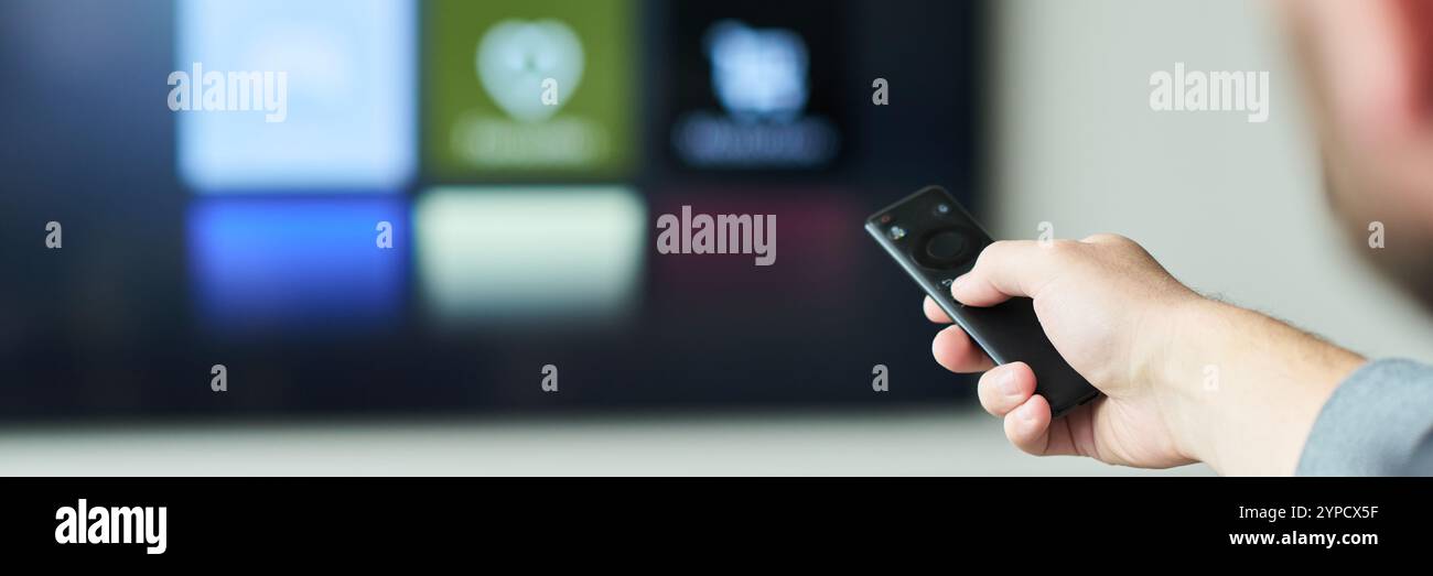 Hand gripping TV remote control in front of blurred television screen ...