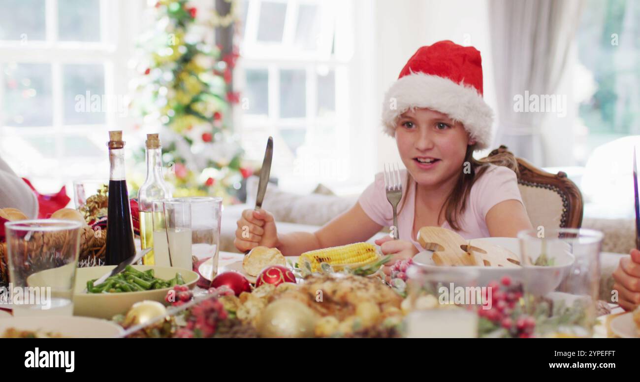 Caucasian girl in santa hat holding fork and knife while sitting on dining table and ready to ...