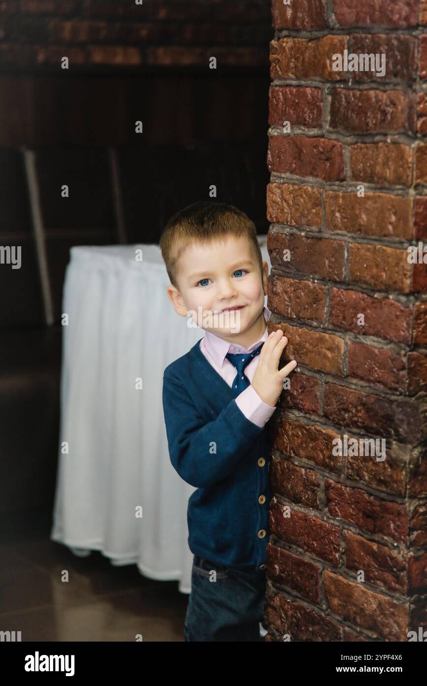 Smiling boy wearing formal attire hiding behind a brick wall during a ...
