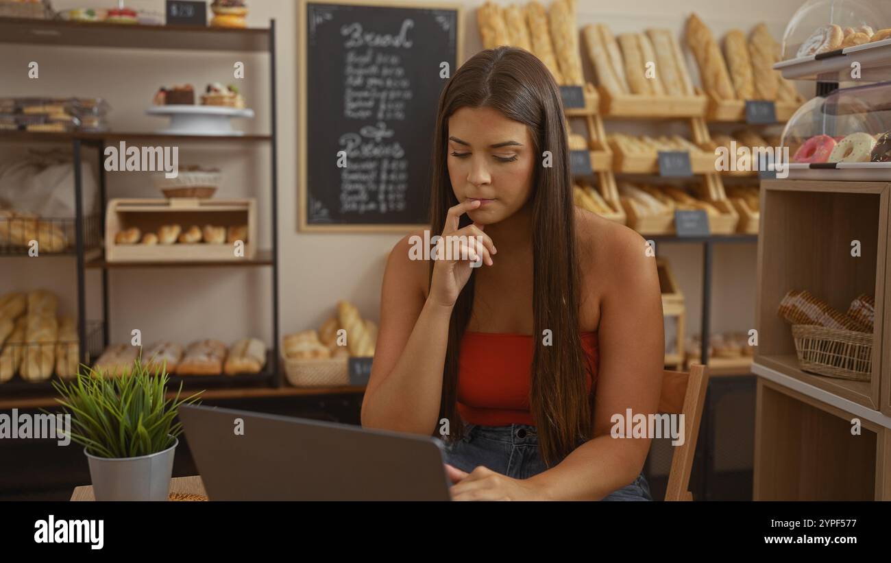 Young woman thinking while using a laptop in a bakery shop with shelves ...