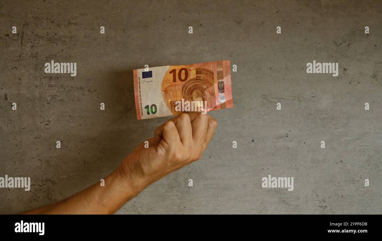 Hand holding a ten-euro banknote against a rough concrete wall surface ...