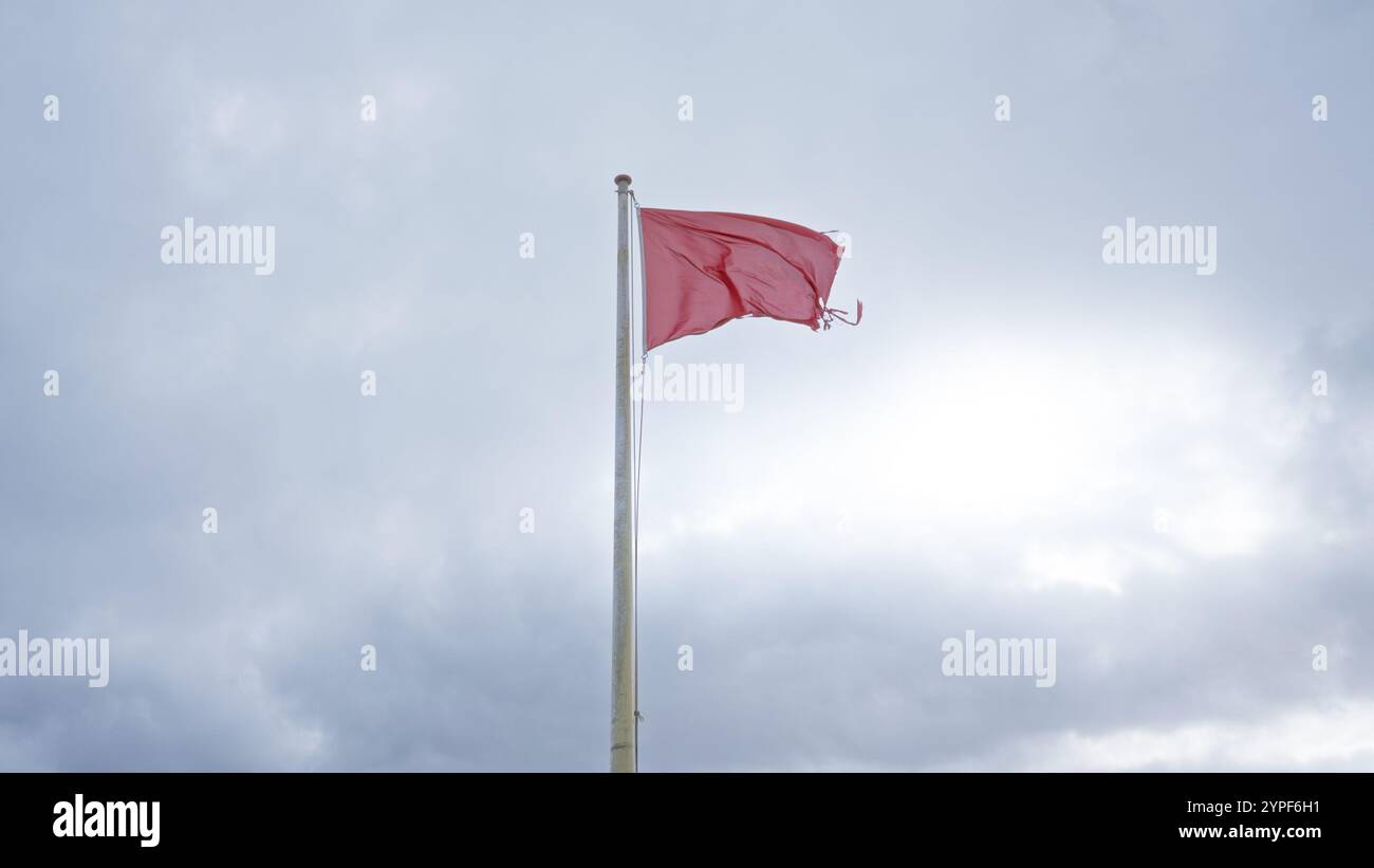 Tattered red flag waving on a pole against a cloudy sky in an outdoor ...