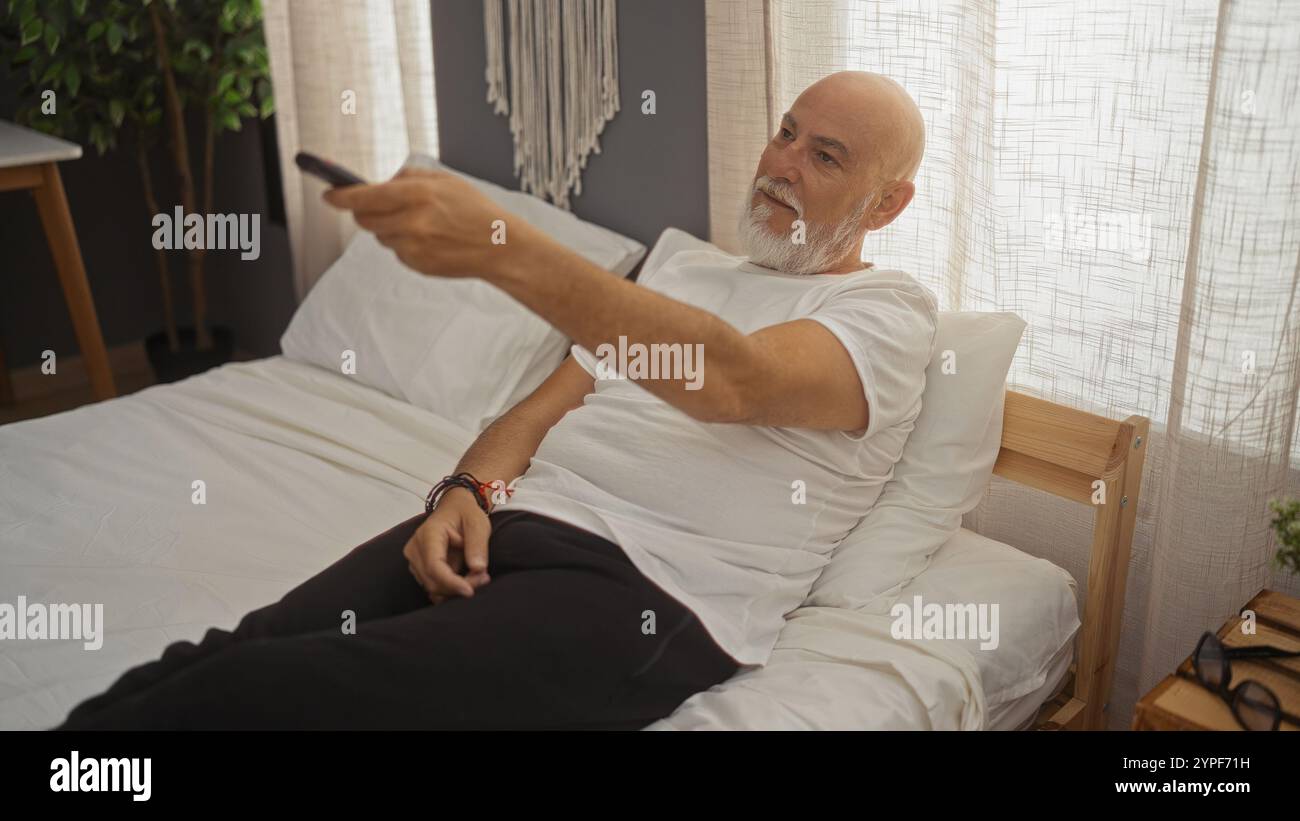 Man relaxing on a bed in a cozy bedroom, holding a remote control, grey ...