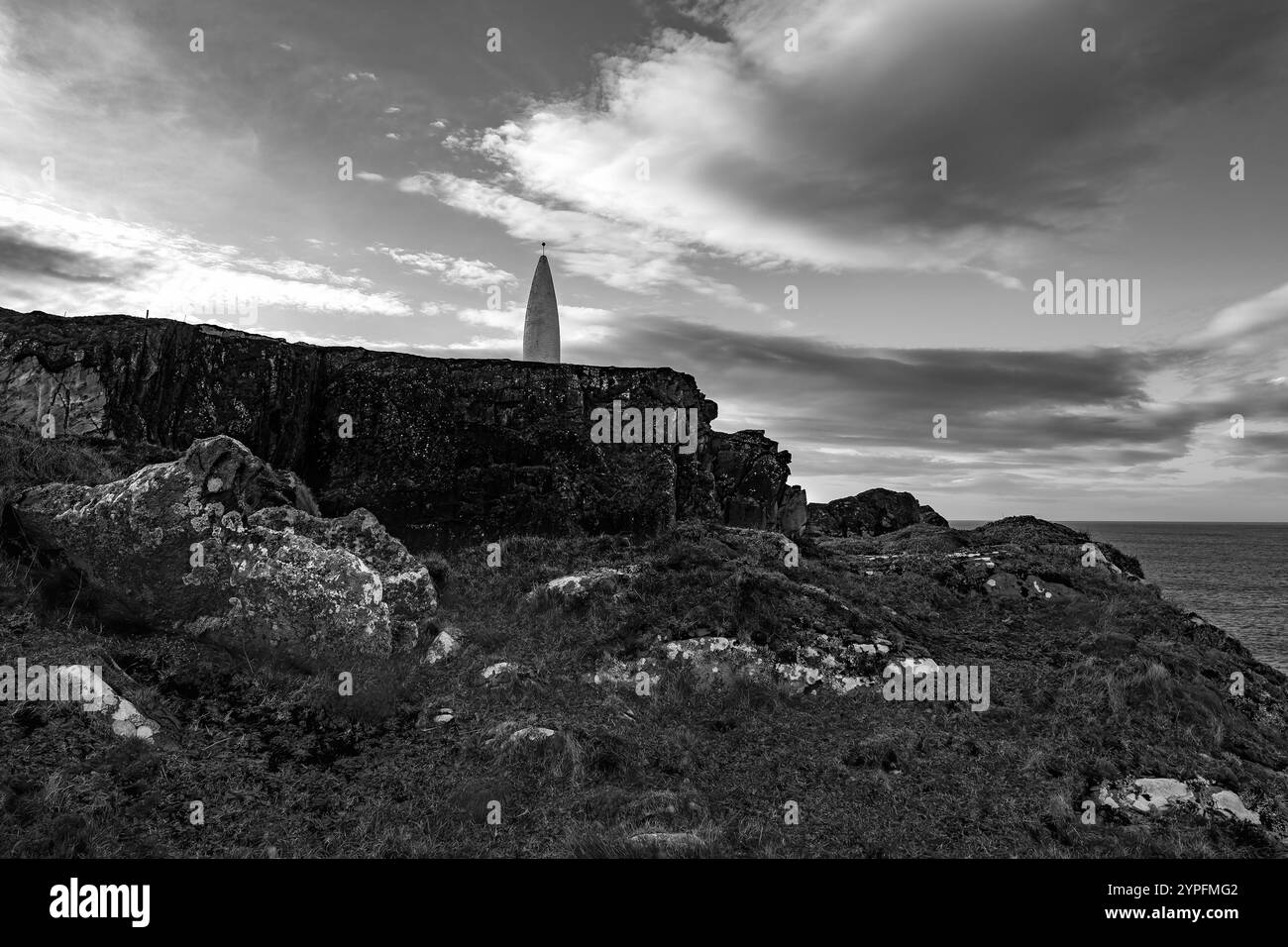 Grey rocks and grass cover a hillside. A slender, light-colored tower ...