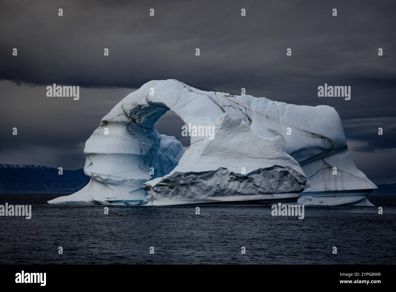 Dark and stormy skies over a blue iceberg that has been carved into an ...