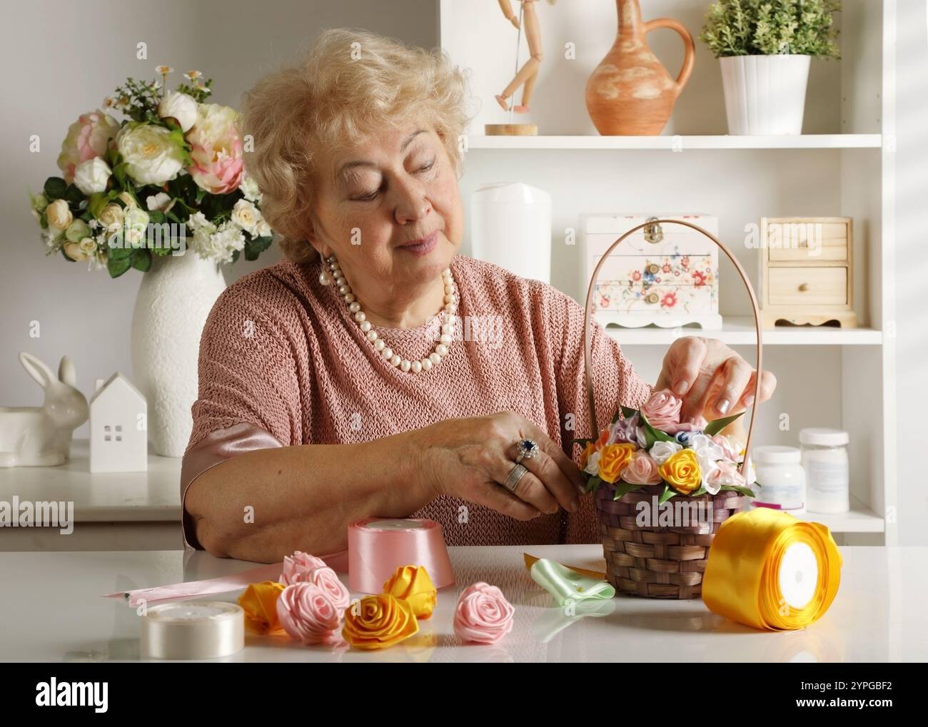 An old woman makes a basket of flowers from ribbons sitting at a table ...