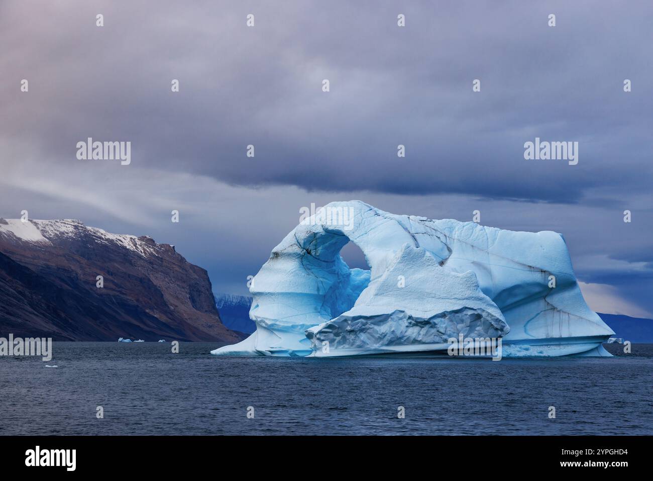 Dark and stormy skies over a blue iceberg that has been carved into an ...