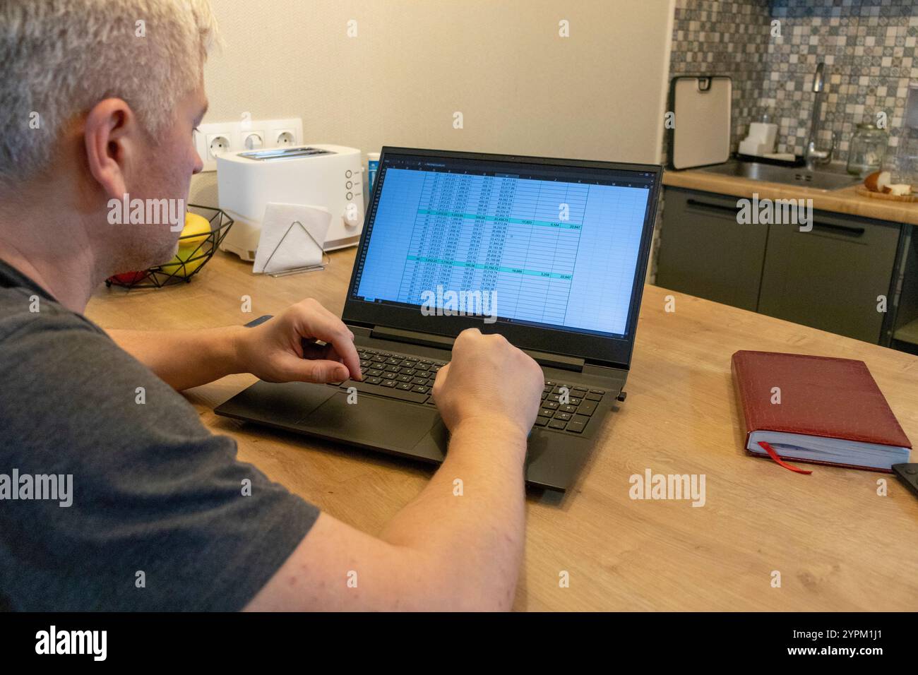 Man working on laptop on the kitchen counter top with an excel sheet on ...