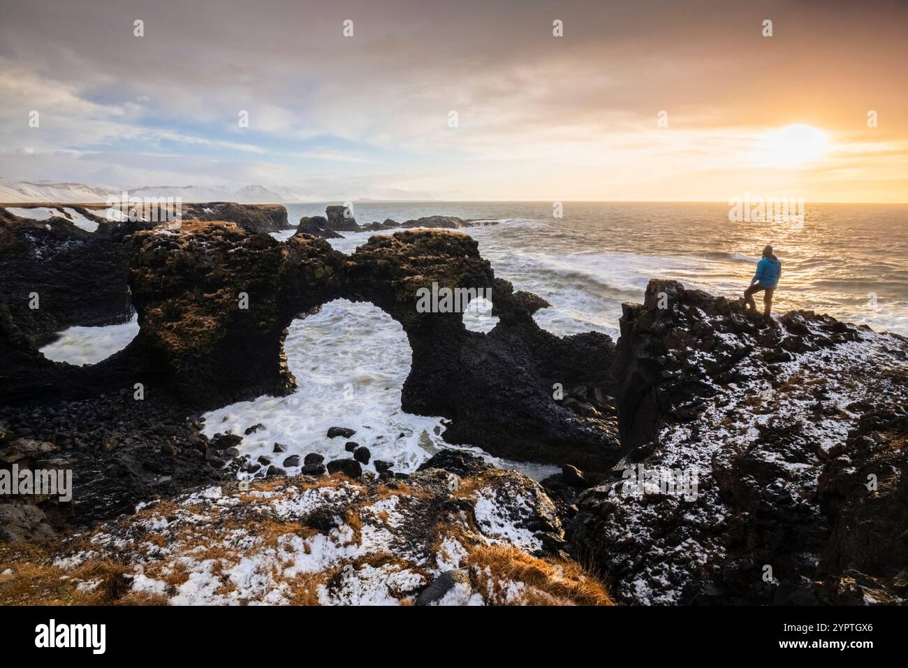 View of a man admiring the Gatklettur rock arch at sunrise in winter. Arnarstapi, Hellnar ...