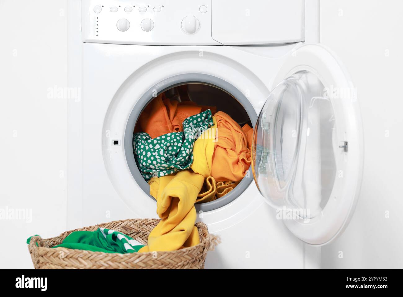 Modern washing machine with clothes and laundry basket near white wall ...
