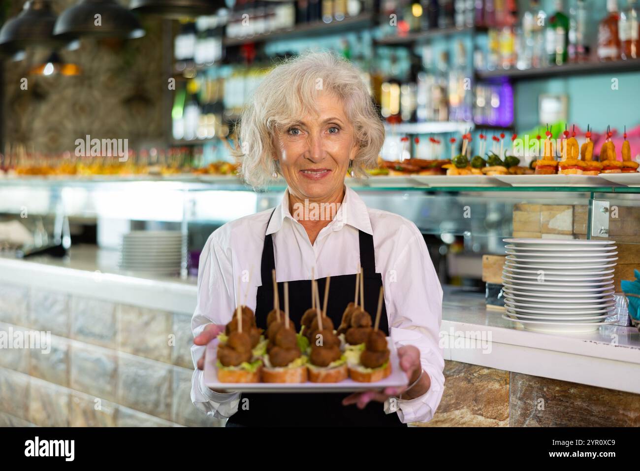 Smiling mature female waiter in apron holding plate with tapas behind a ...