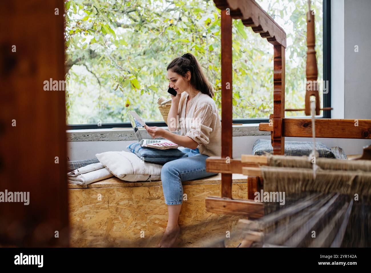 Young businesswoman making a call with customer, sitting by wooden loom ...