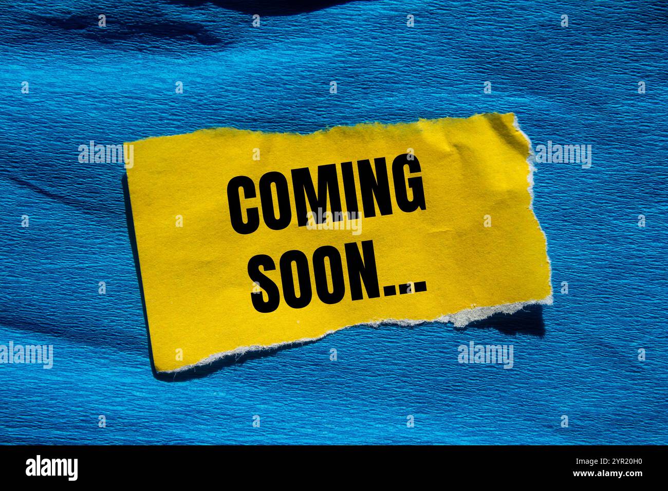 Coming soon message written on ripped torn yellow paper piece with blue ...