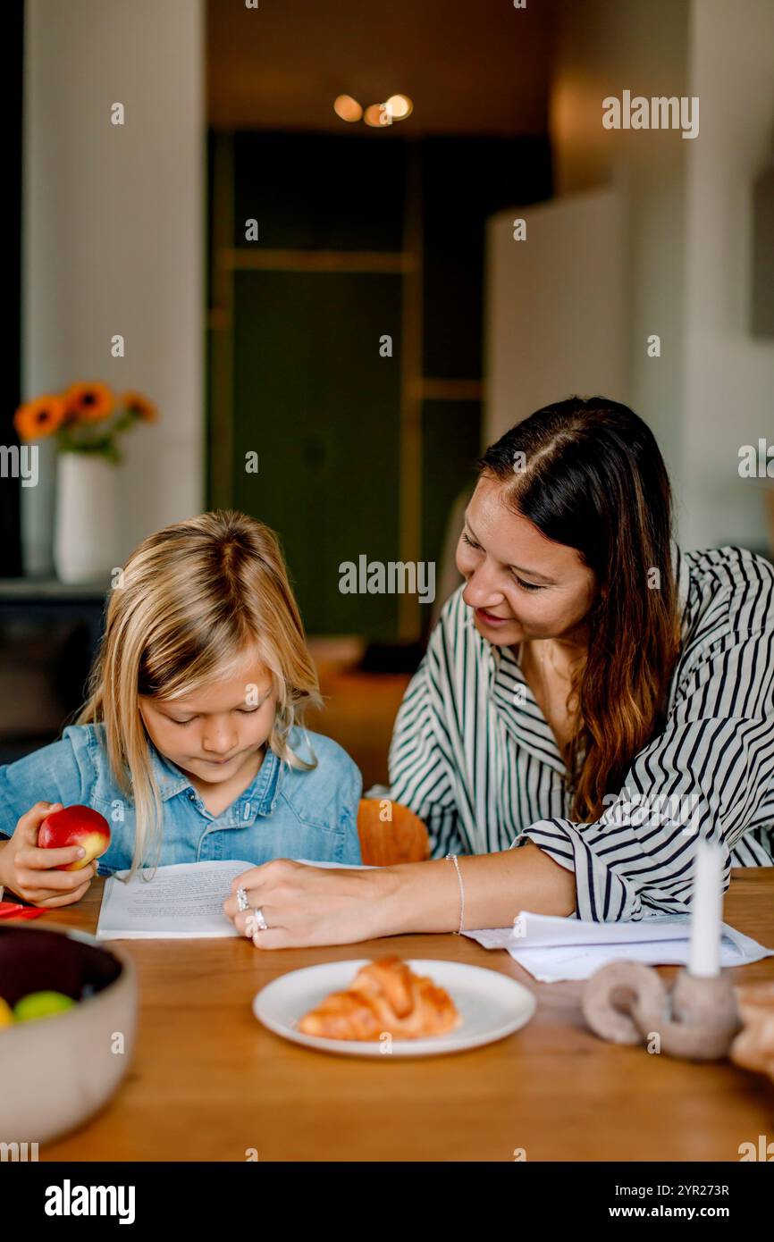 Mother guiding son doing homework while sitting near table at home ...