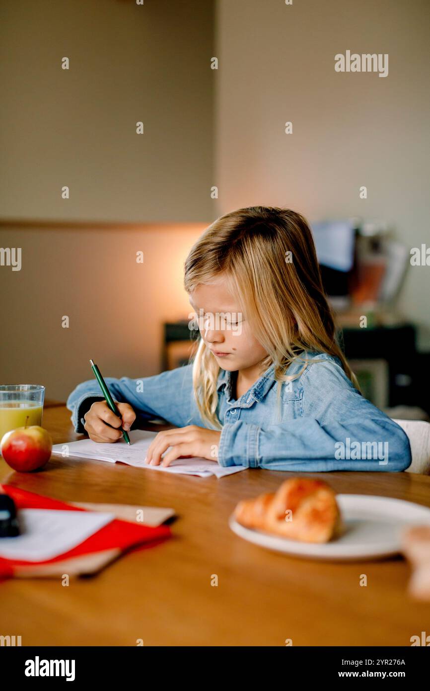 Focused boy with blond long hair writing on book while dong homework at ...