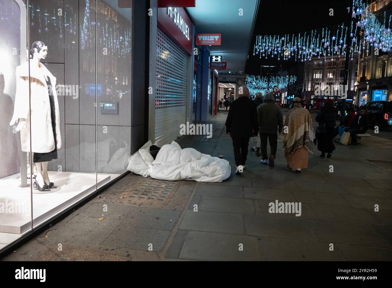 Pedestrian pass by a homeless man asleep in a duvet outside a shop ...