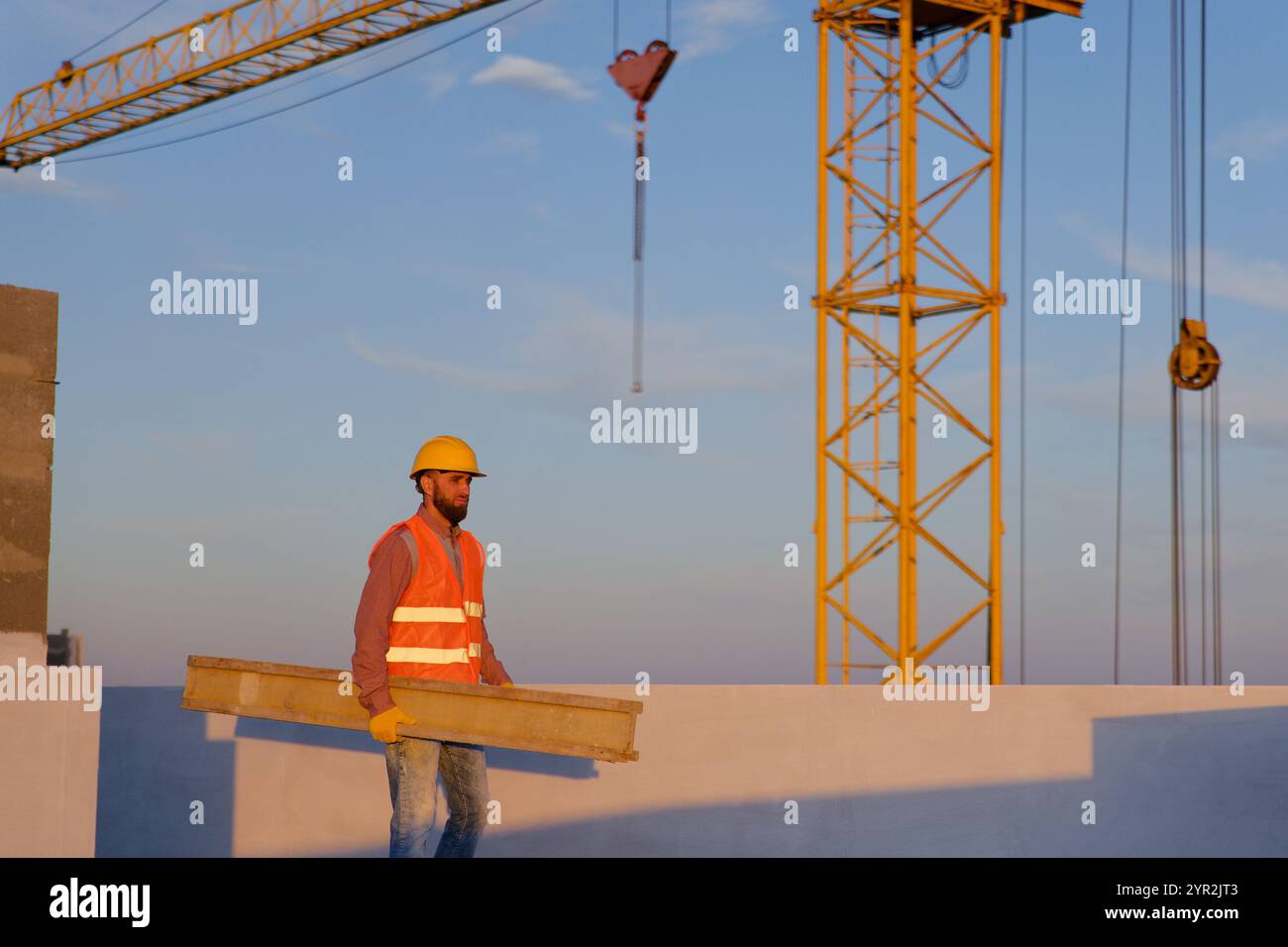 Construction worker carrying a wooden plank on a rooftop at sunset ...