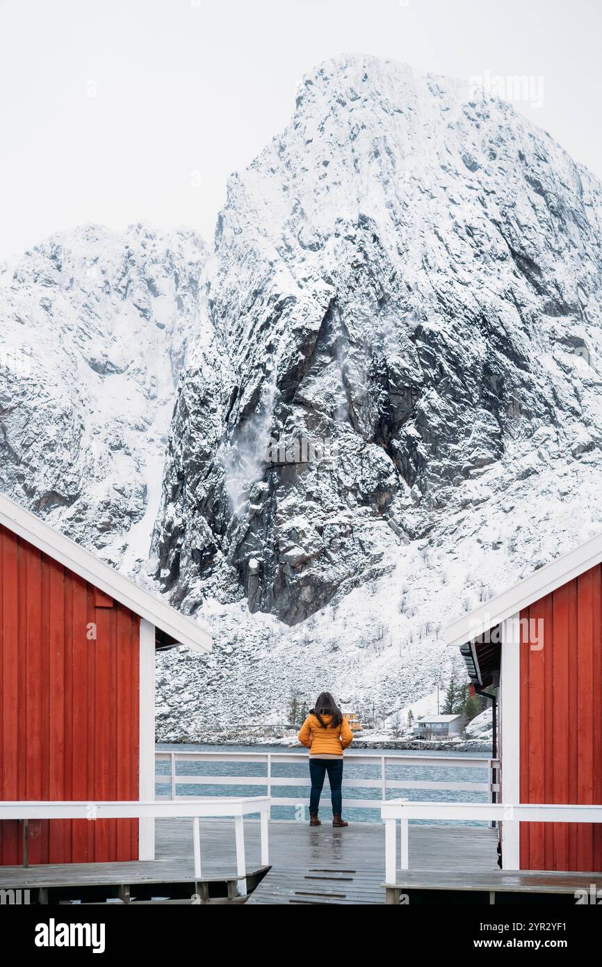 Tourist admiring snowy mountains and red rorbuer in lofoten islands ...