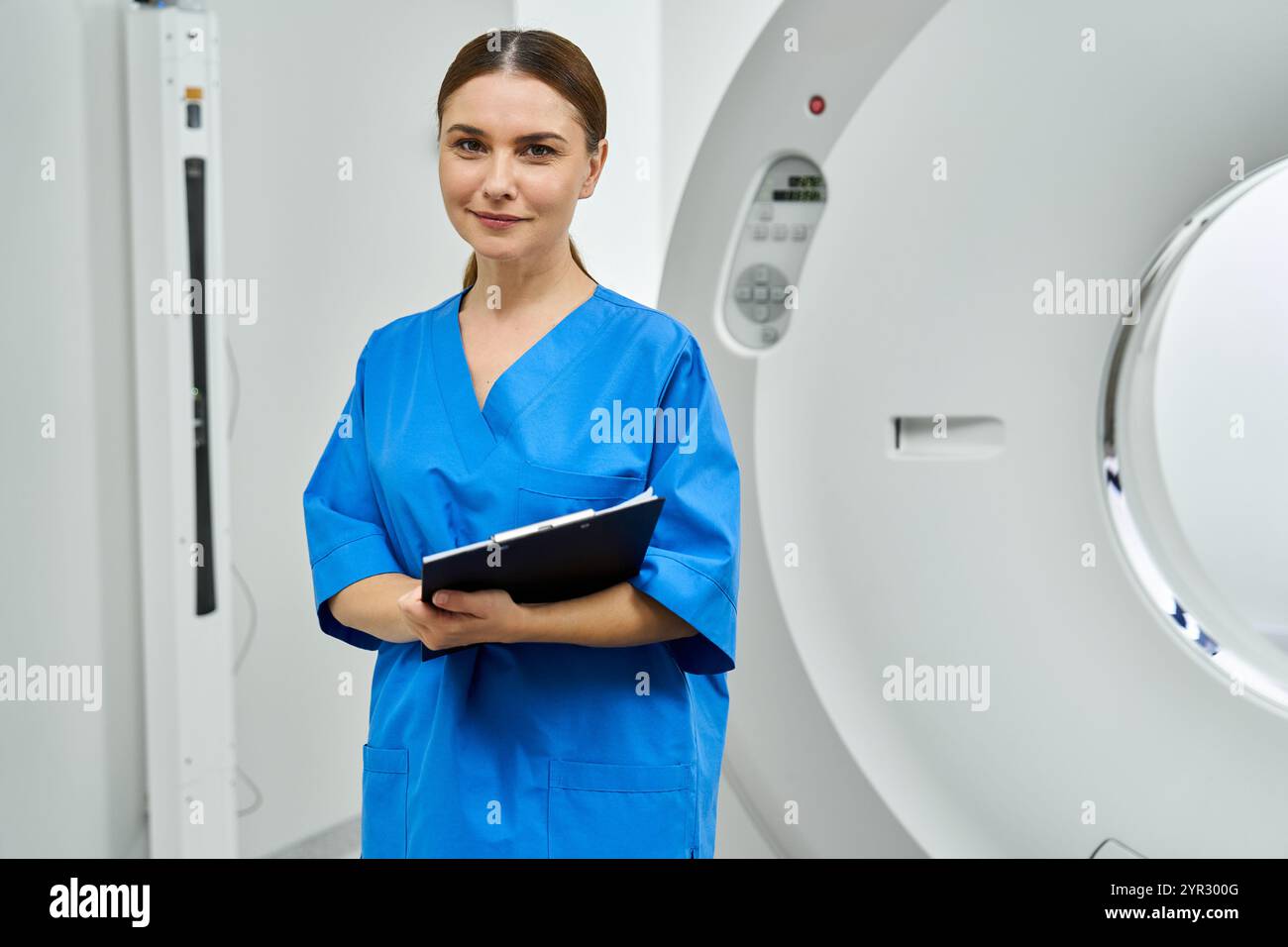 Attractive female doctor in blue scrubs stands confidently next to MRI ...