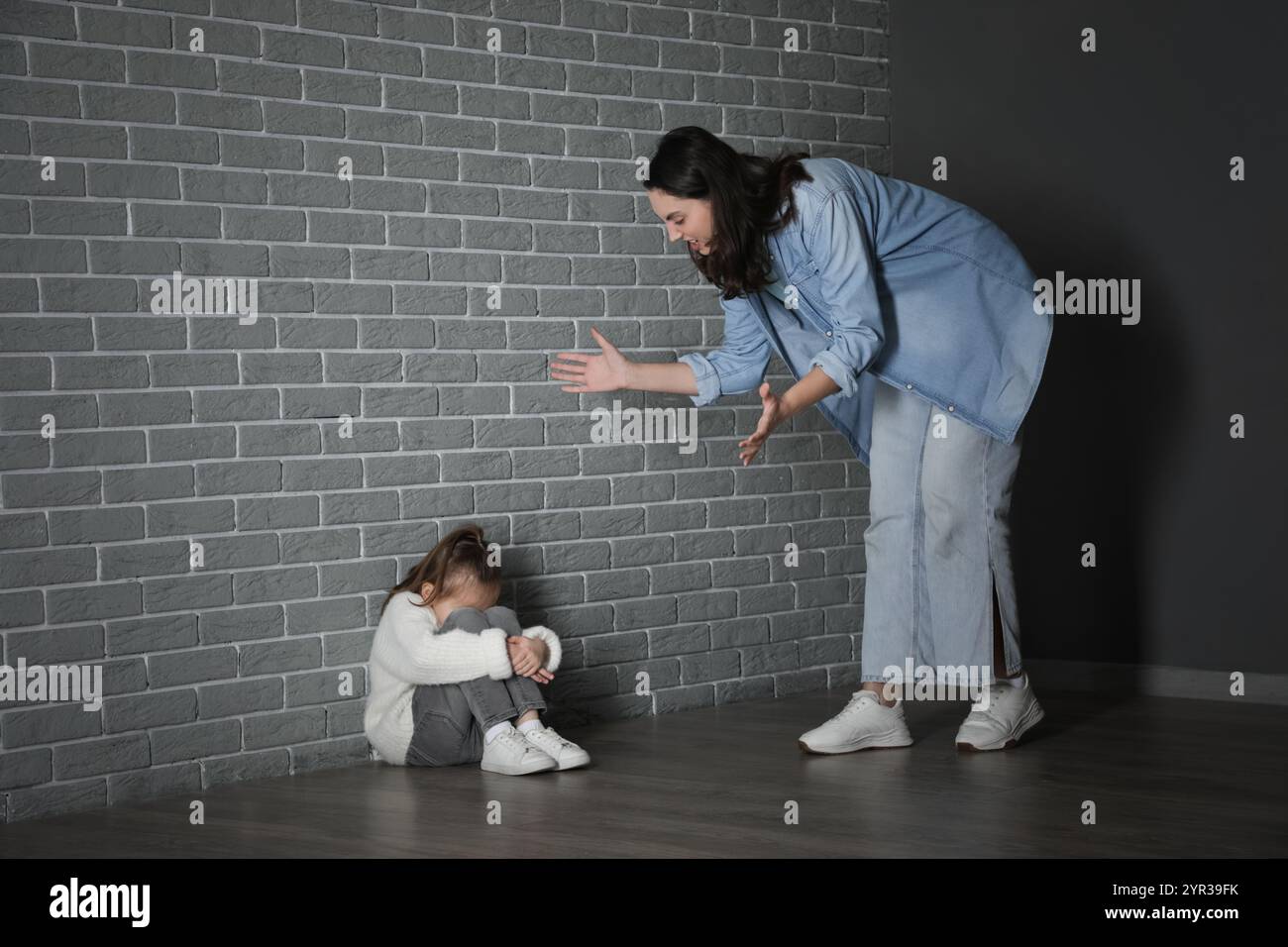 Scared little girl with her angry mother sitting near grey brick wall ...