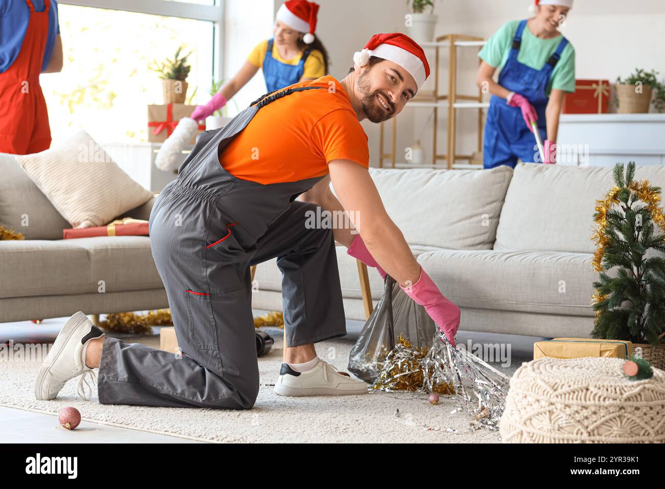 Male janitor with Santa hat putting tinsel into garbage bag in room ...