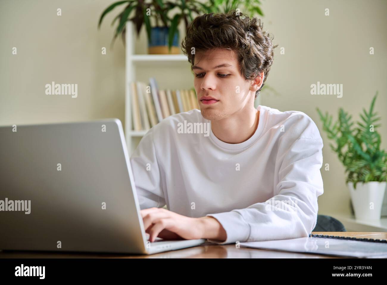 Young male college student sitting at desk at home typing on laptop ...