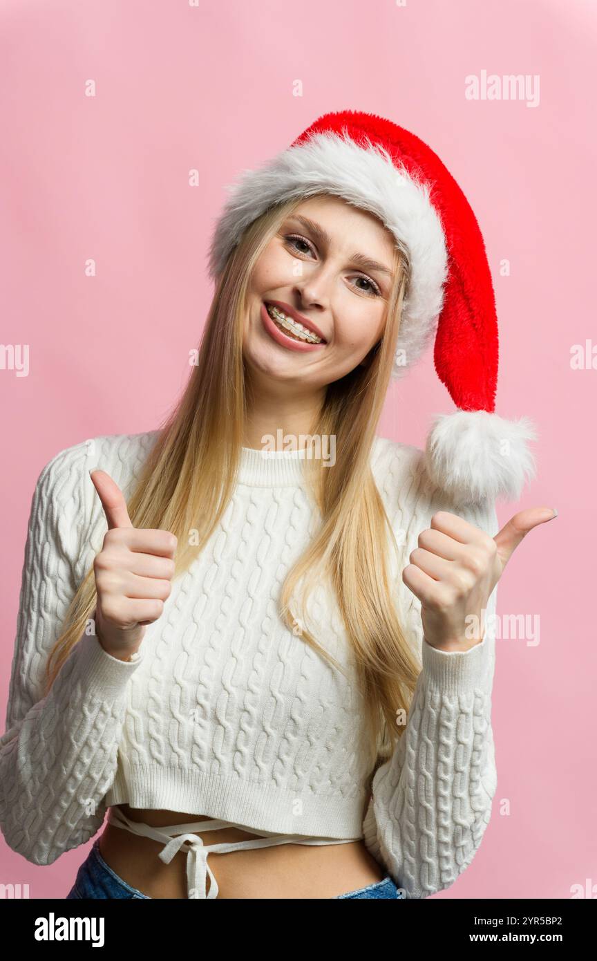 A smiling young woman poses with thumbs up, wearing a Santa hat against ...