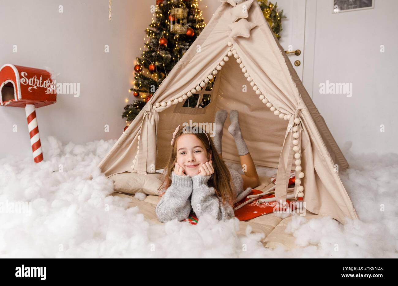 A charming girl, nestled in a cozy Christmas teepee, smiles happily ...