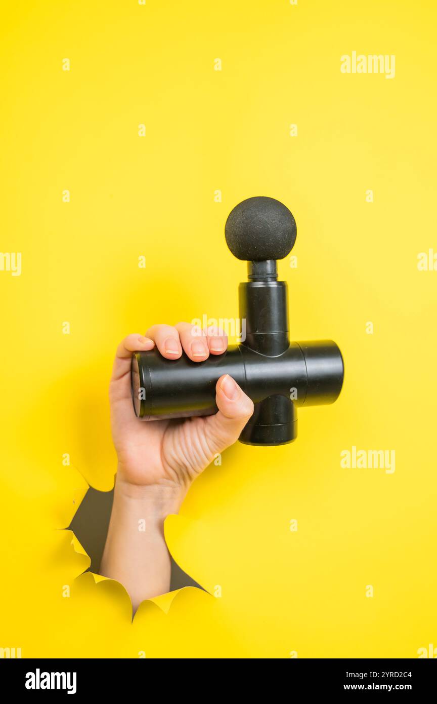 Female hand sticking out with massage gun from yellow paper background ...