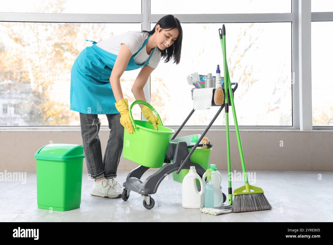 Female janitor with bucket and trolley of cleaning supplies in room ...