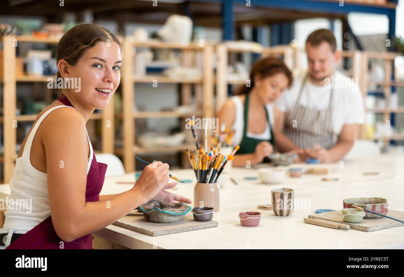 Girl in apron designing handmade clay plate with brush in taller room ...