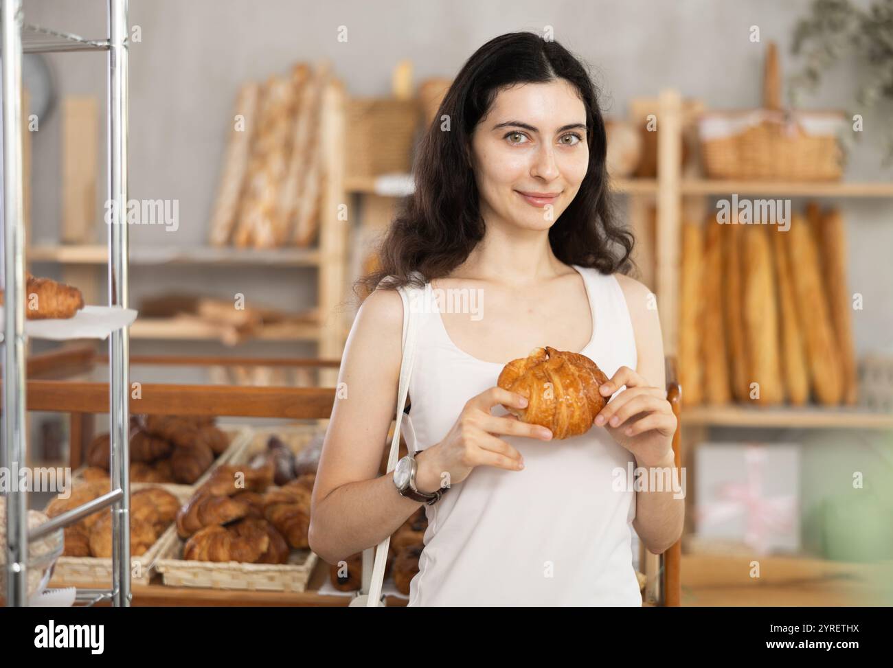 Young female customer stands with croissant in hands near window of ...