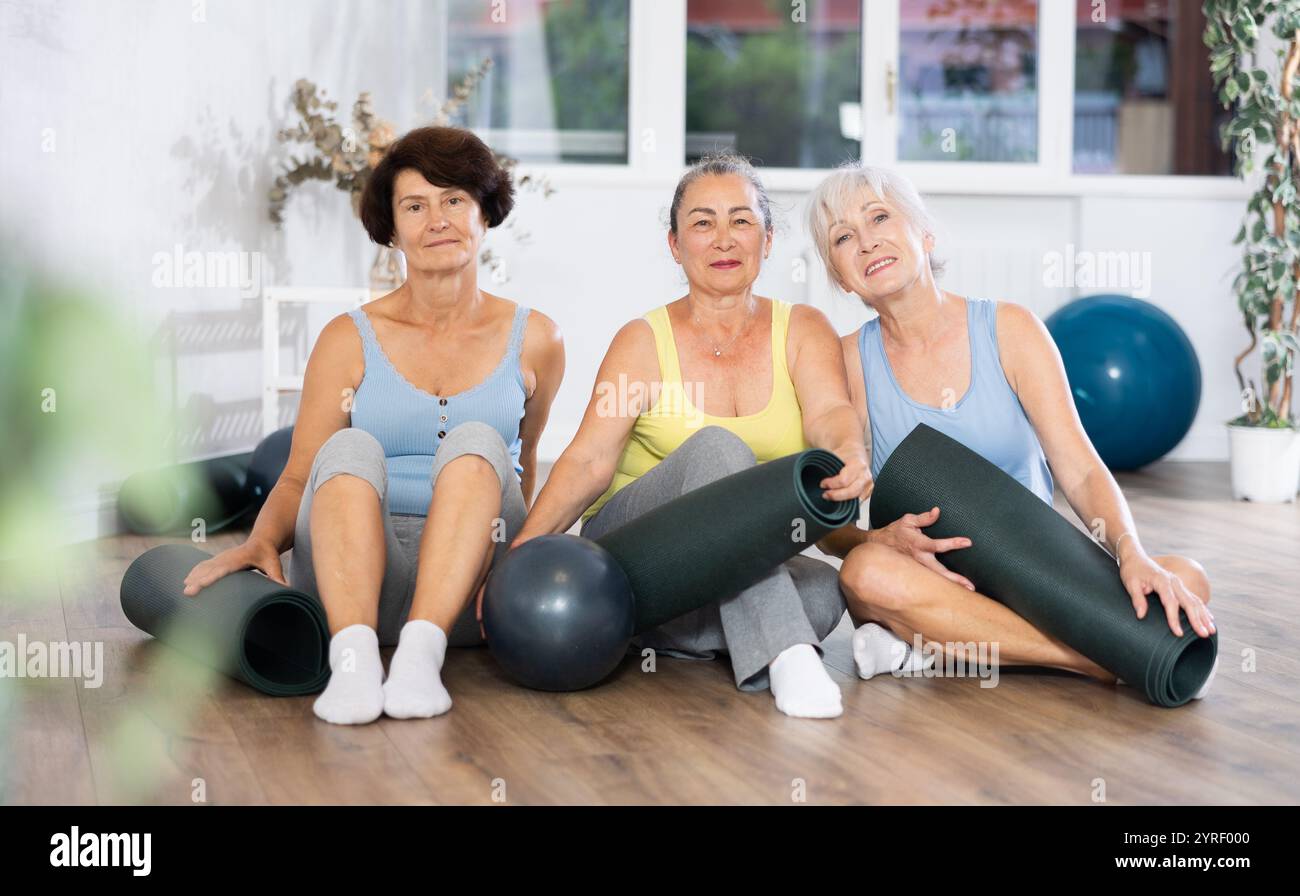 Mature women posing in seated position holding yoga rugs in fitness ...