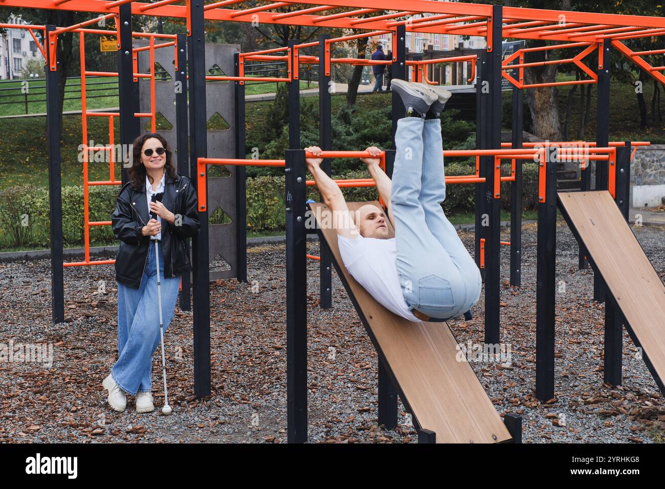 Two friends enjoy a day at the park gym. A blind man hangs from bars ...