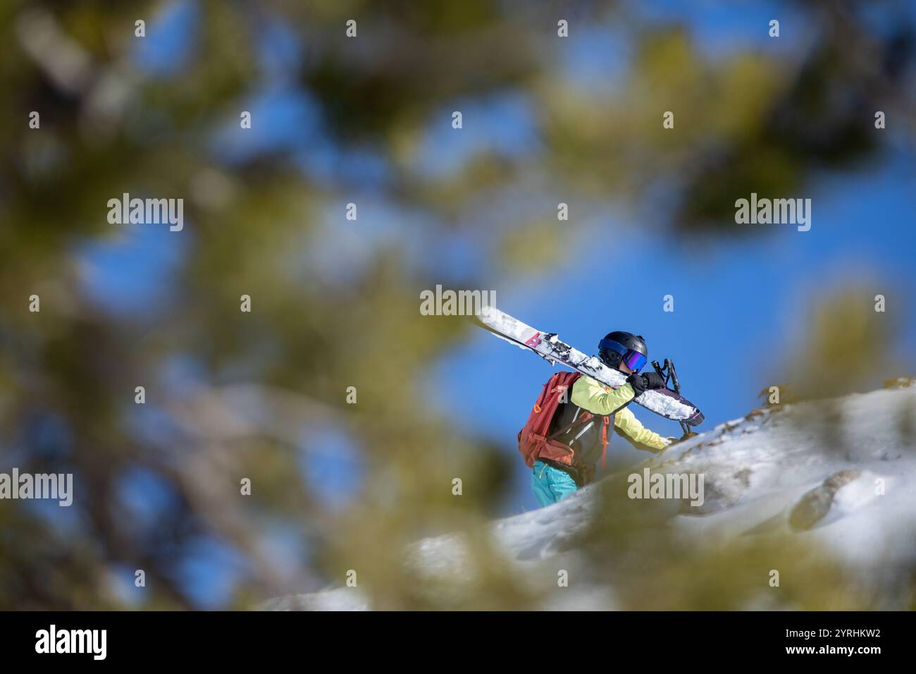 A woman in bright ski gear hikes up a snowy mountain, carrying skis on her shoulder under a ...