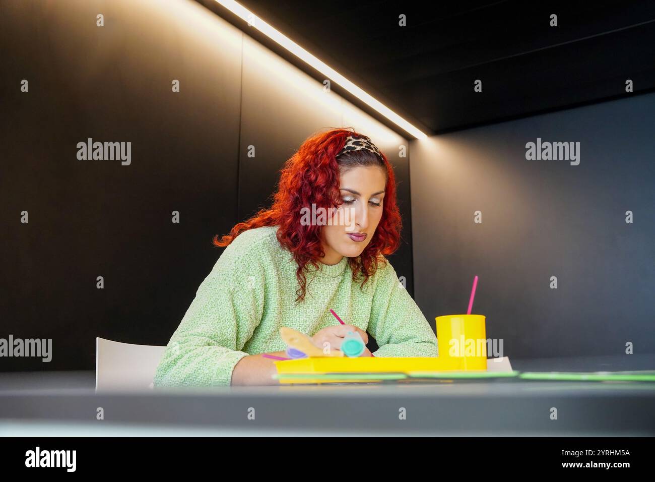 A trans woman with red hair works intently at a desk, surrounded by art ...