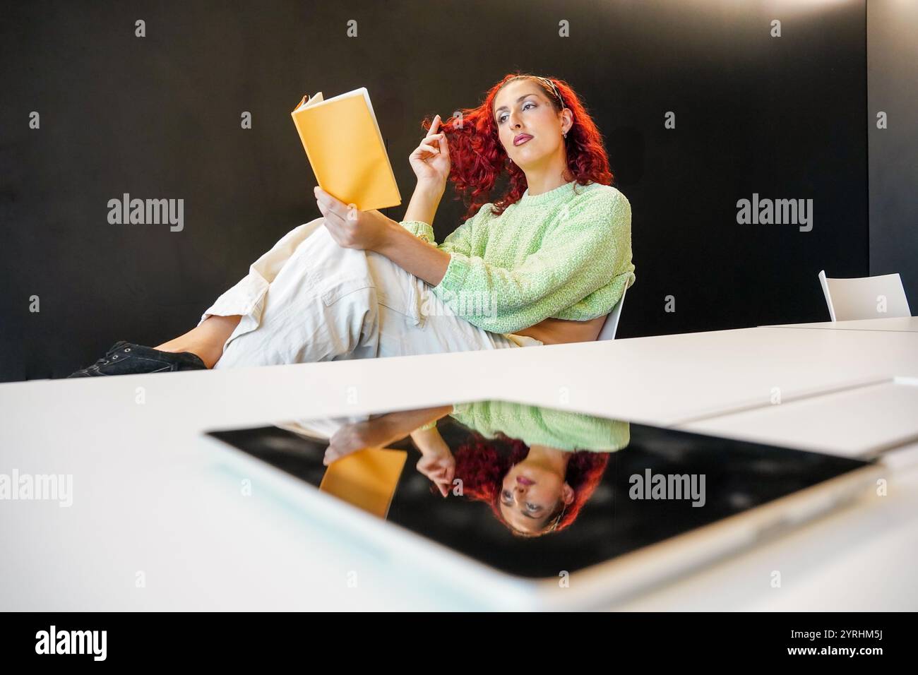 A trans woman with red hair relaxes in a modern work space, reflecting ...