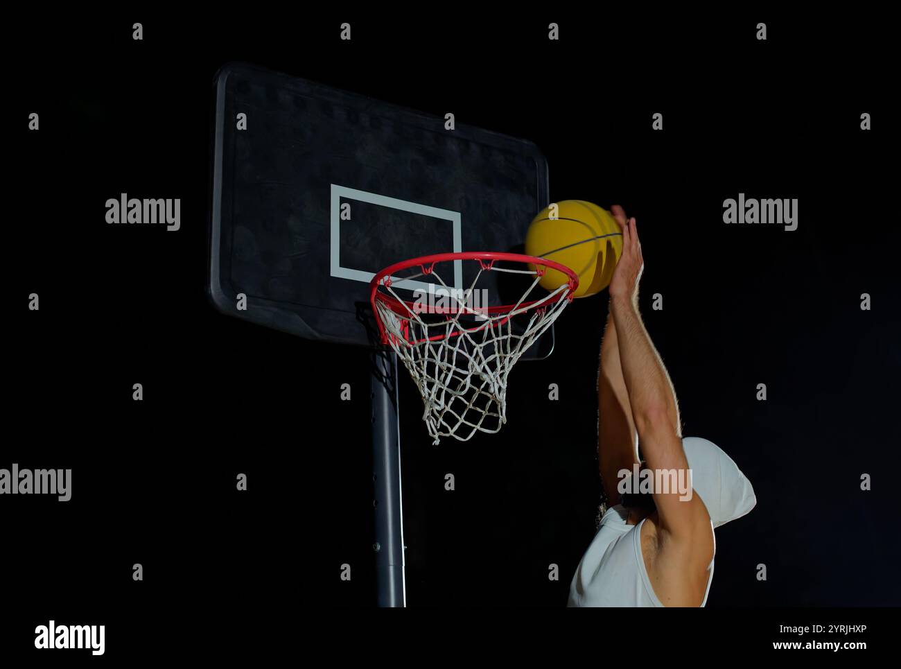 A man dressed in a white tank top and shorts jumps high to score a slam dunk on an outdoor ...