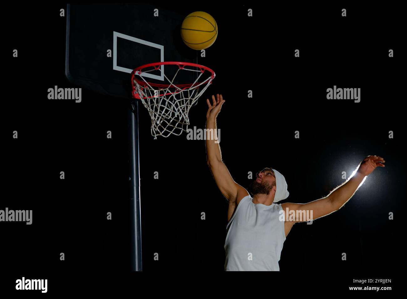 A man dressed in a white tank top and shorts jumps high to score a slam dunk on an outdoor ...