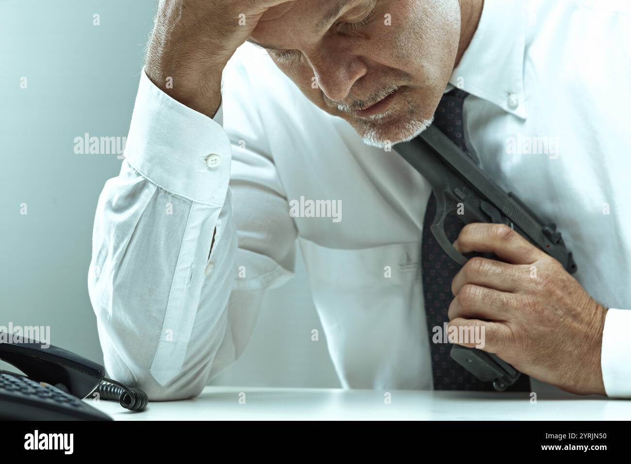 Stressed mature businessman sitting at his desk, gripping a gun under ...