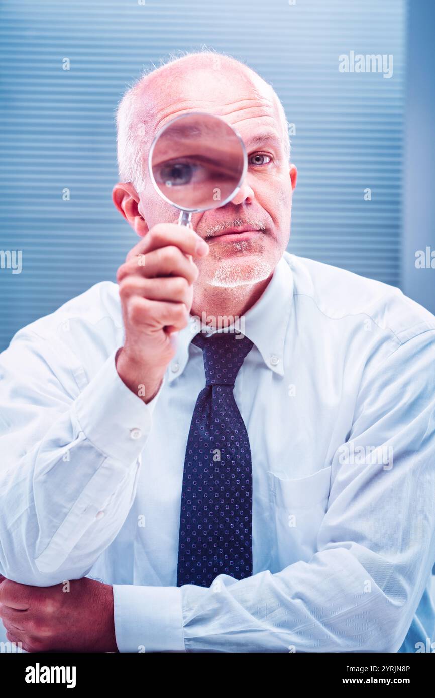 Mature businessman holding a magnifying glass over his eye, looking ...